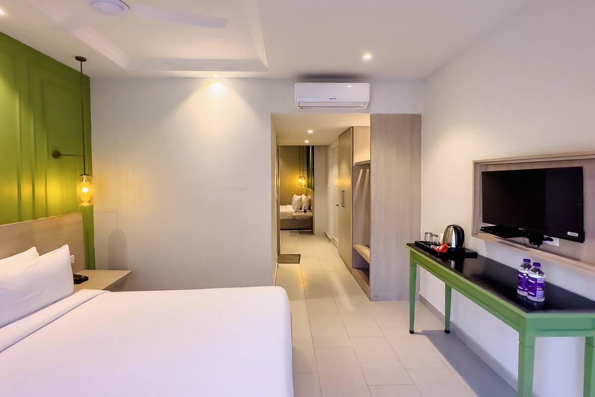 Quarto de hotel com cama, escrivaninha com televisão, ventoinha no teto e corredor que dá para um banheiro.