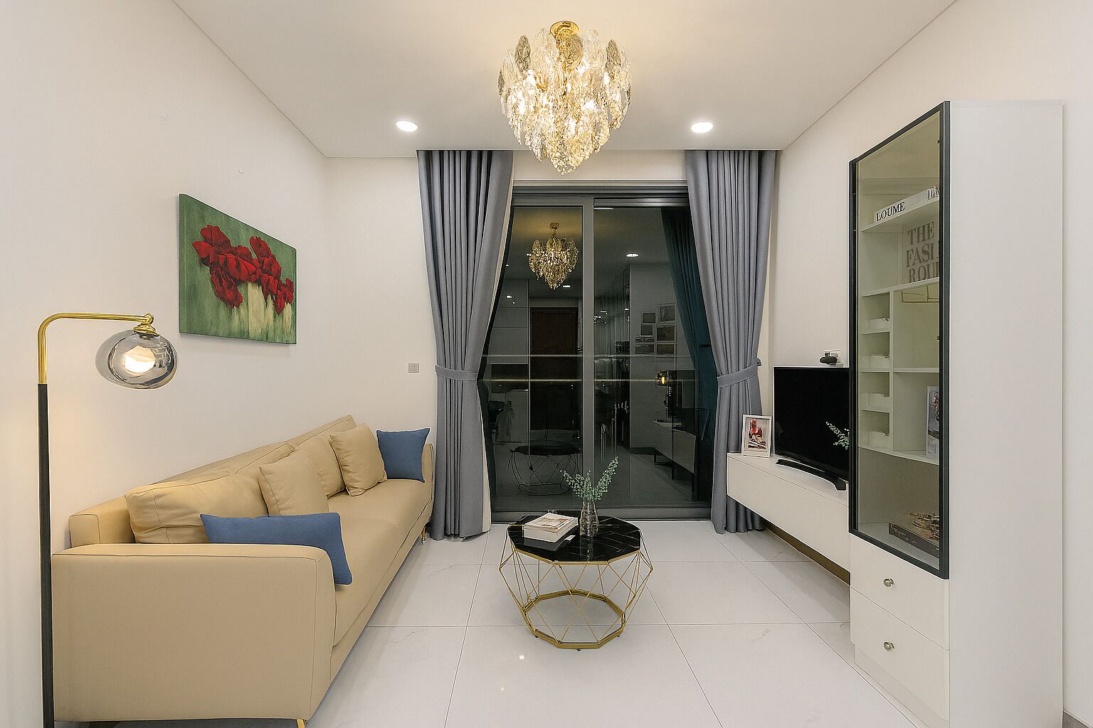 Uma sala de estar moderna com um sofá bege, uma mesa de centro de vidro, um candelabro e uma TV em um armário branco.