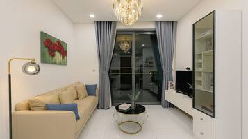 Uma sala de estar moderna com um sofá bege, uma mesa de centro de vidro, um candelabro e uma TV em um armário branco.