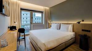 Quarto de hotel moderno com uma cama grande, uma escrivaninha e uma cadeira. Há uma janela com cortinas, um abajur e uma pequena mesa com um telefone.