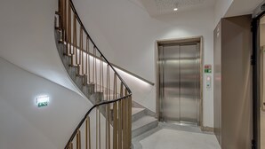 Um elevador moderno com acabamento metálico, uma escada curva com guarda-corpos de madeira e um sinal de saída de emergência fixado na parede.