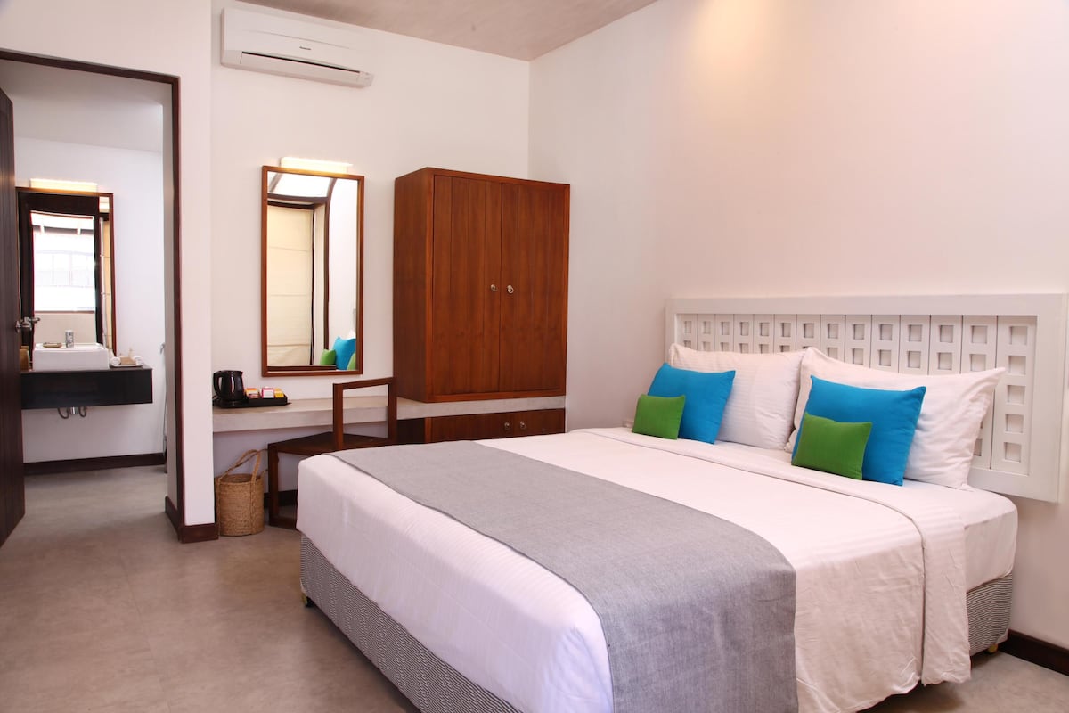 Quarto de hotel com uma cama, um guarda-roupa de madeira, uma escrivaninha com espelho e cabeceira branca.