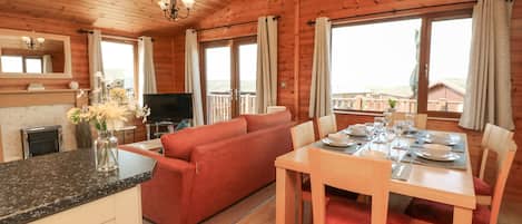Interior de uma cabana de madeira com sala de jantar, sofá, TV e janelas amplas.