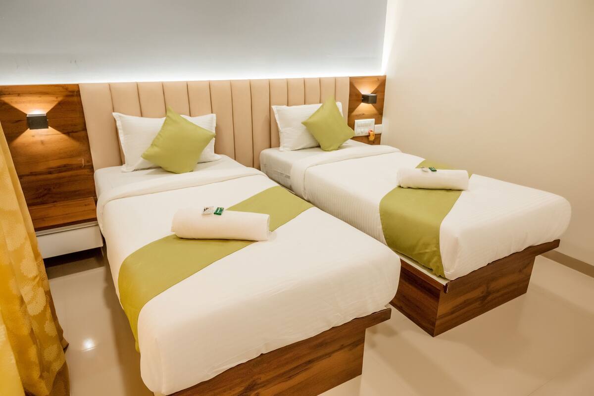 Quarto de hotel com duas camas de solteiro, cada uma com cabeceira de madeira, roupa de cama branca e almofadas decorativas verdes. Há mesas de cabeceira com abajures e um painel de parede de madeira.