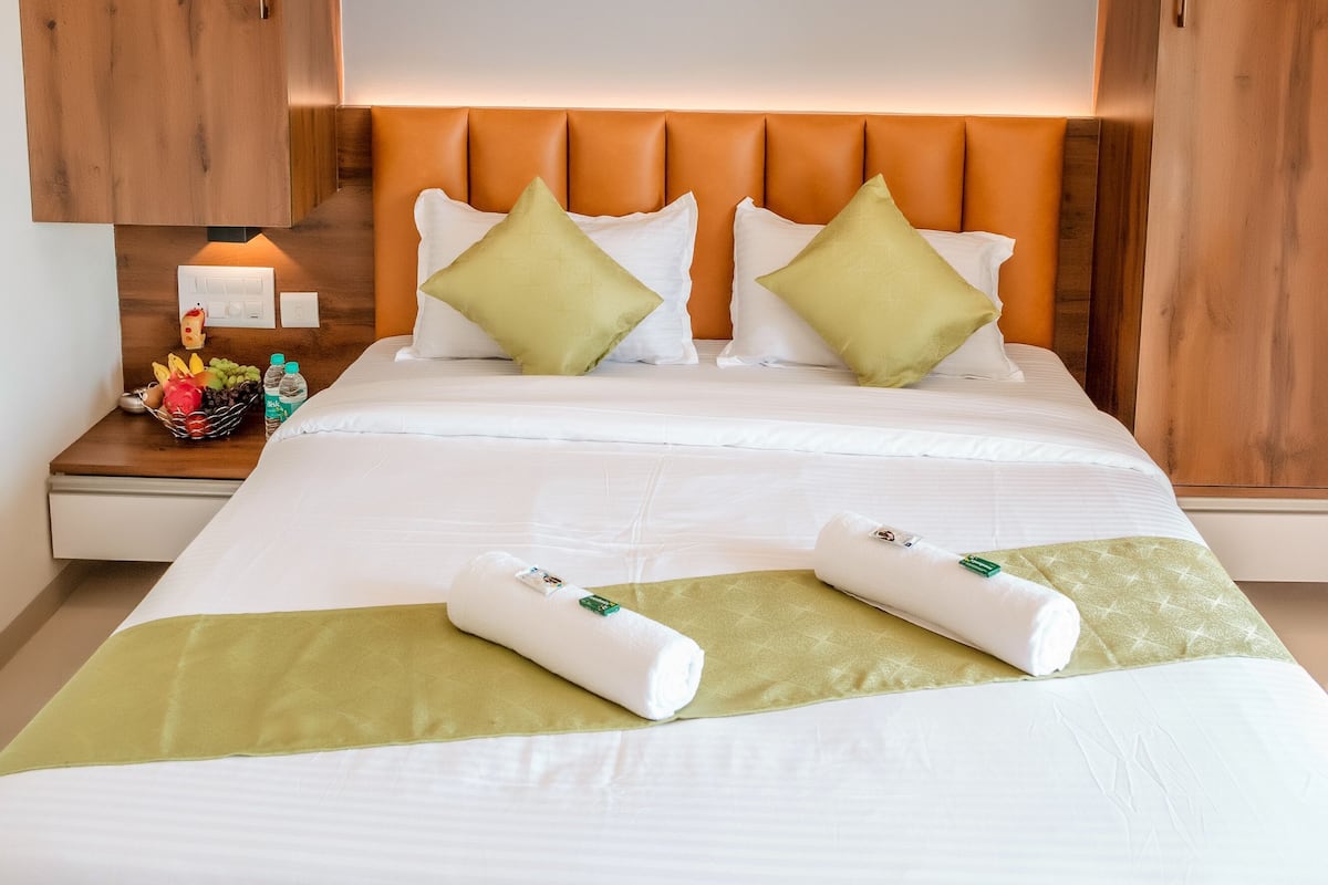 Quarto de hotel com cama, cabeceira de madeira, roupa de cama branca e mesa de cabeceira com luminária e cesta de frutas.