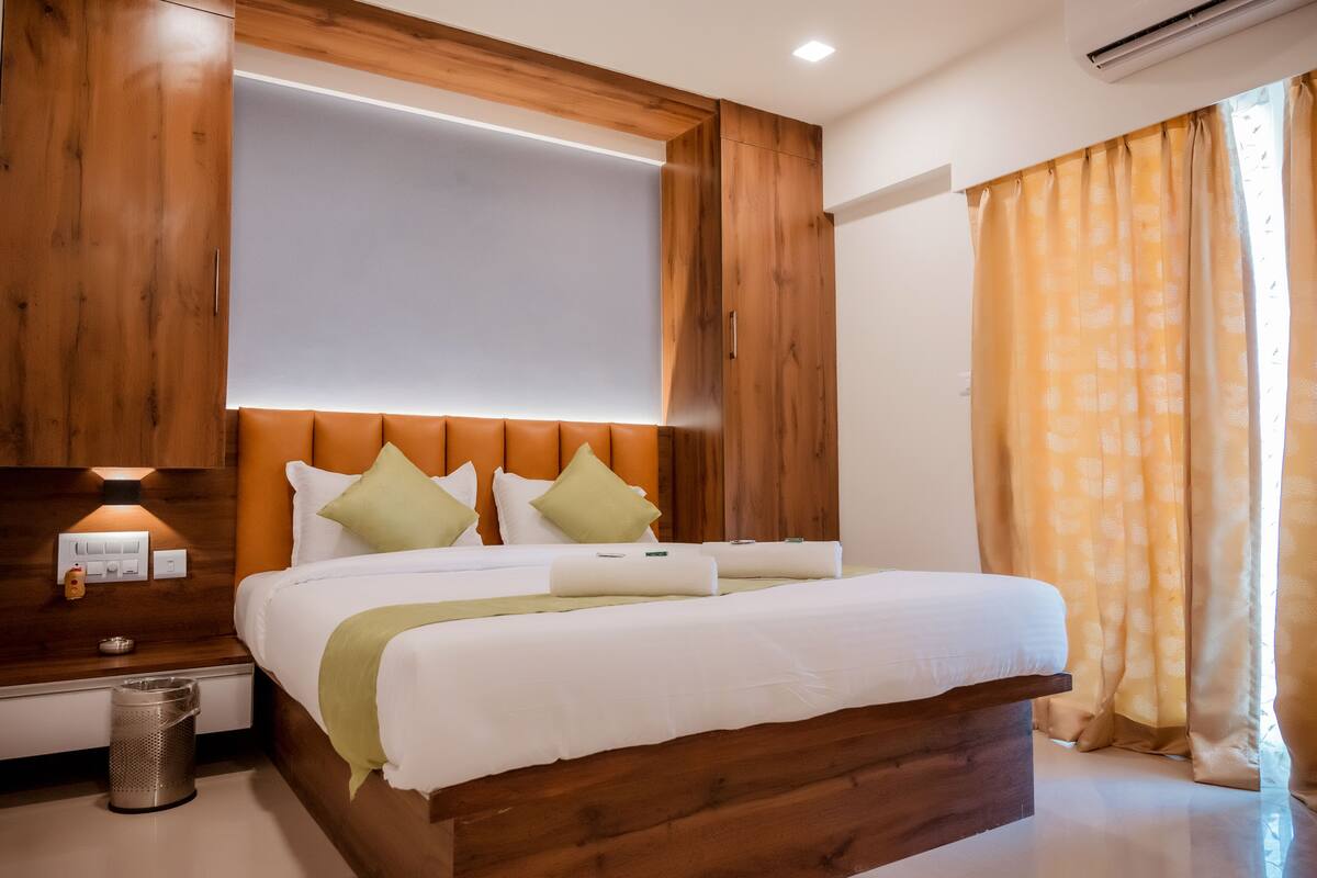 Quarto de hotel com cabeceira de madeira, cama com roupa de cama branca e almofadas verdes, mesa de cabeceira com abajur e janela com cortinas amarelas.