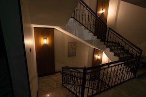 Uma escadaria com corrimão de ferro negro trabalhado, uma porta de madeira na base e luminárias fixadas nas paredes em ambos os lados.