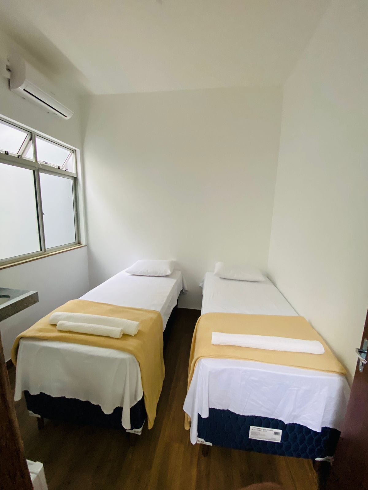 Quarto pequeno e limpo com duas camas de solteiro, uma janela e piso de madeira.