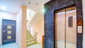 Um saguão de hotel com uma escadaria de mármore, um elevador azul escuro e uma porta com a inscrição '101'.