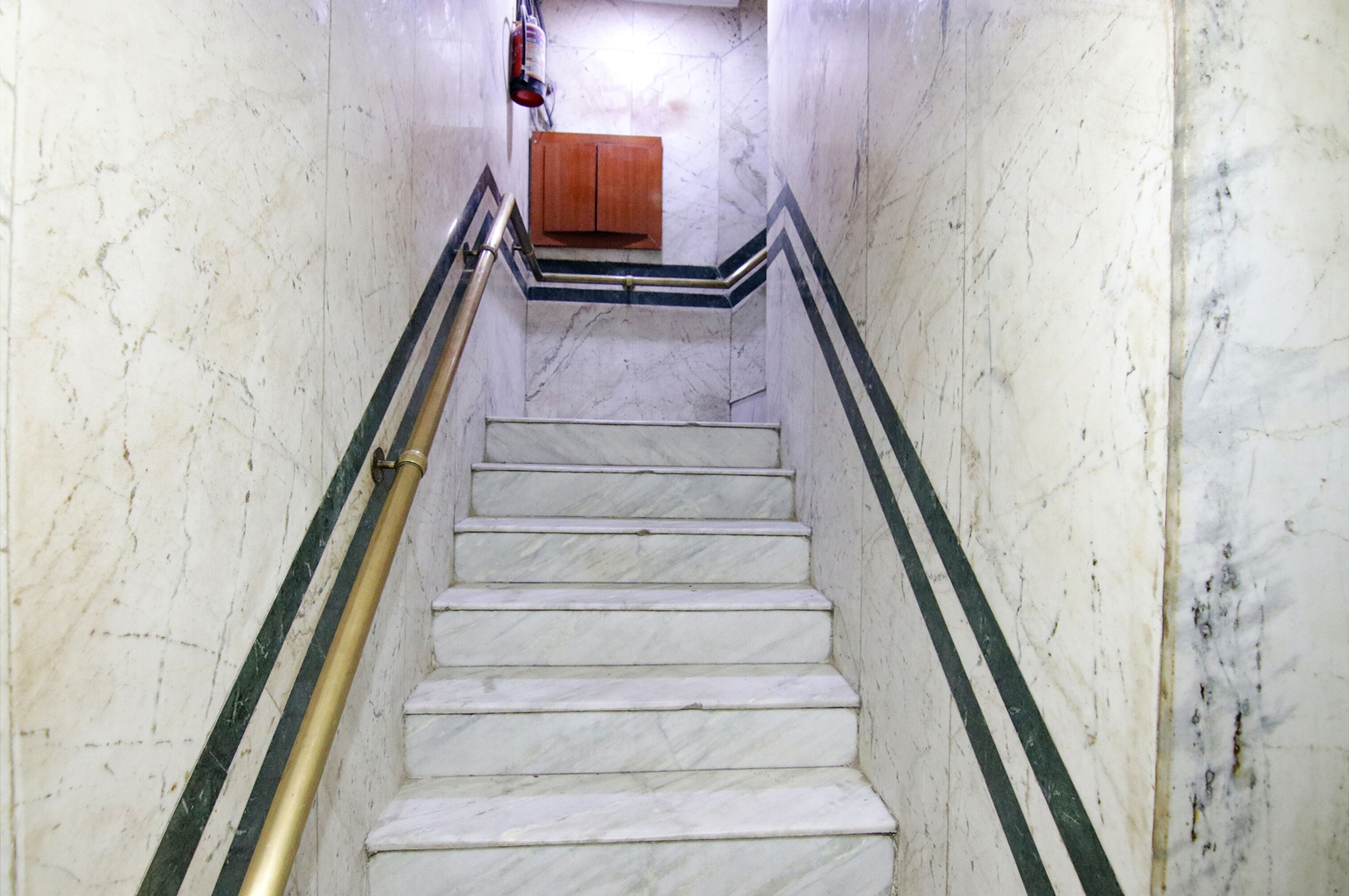 Uma escadaria de mármore com corrimão, um armário de madeira e um extintor de incêndio.
