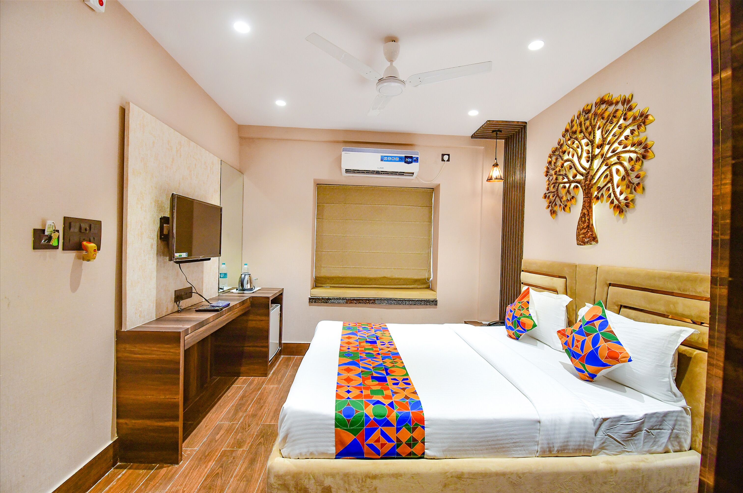 Quarto de hotel com cama, escrivaninha, televisão, ar condicionado e uma árvore decorativa na parede.