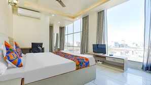 Quarto de hotel moderno com uma cama grande, uma escrivaninha com televisão e vista para a cidade através de grandes janelas.