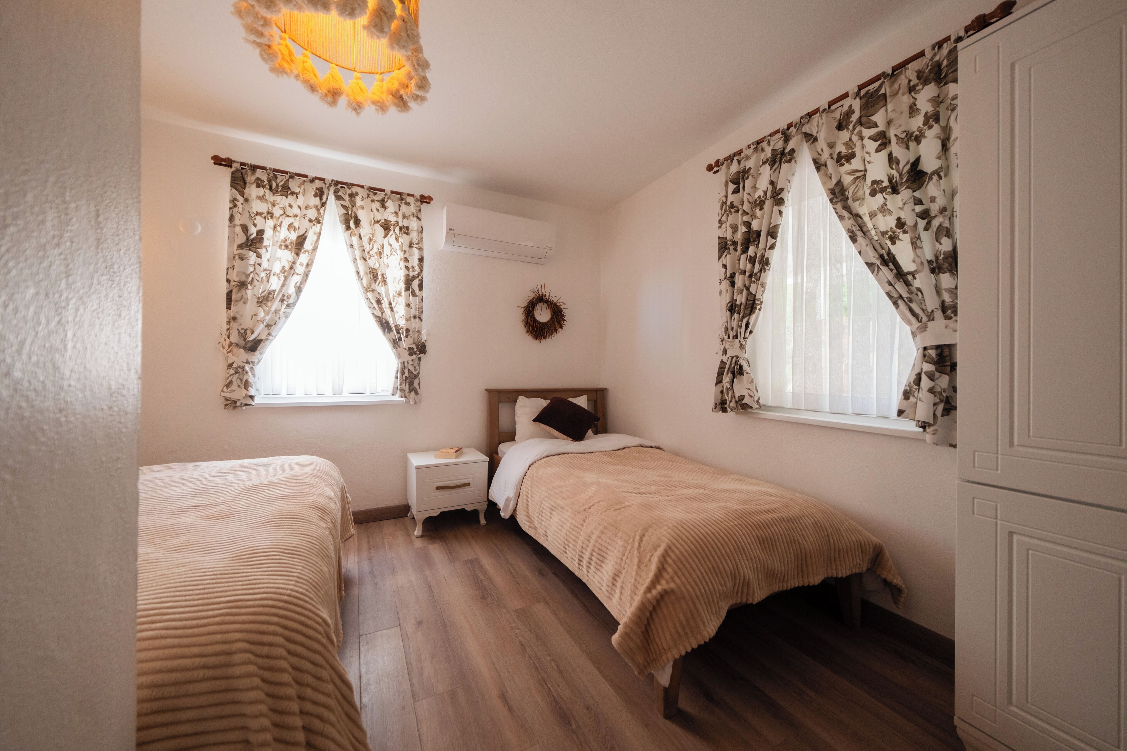 Quarto com duas camas de solteiro, uma mesinha de cabeceira, ar condicionado fixado na parede e janela com cortinas florais.