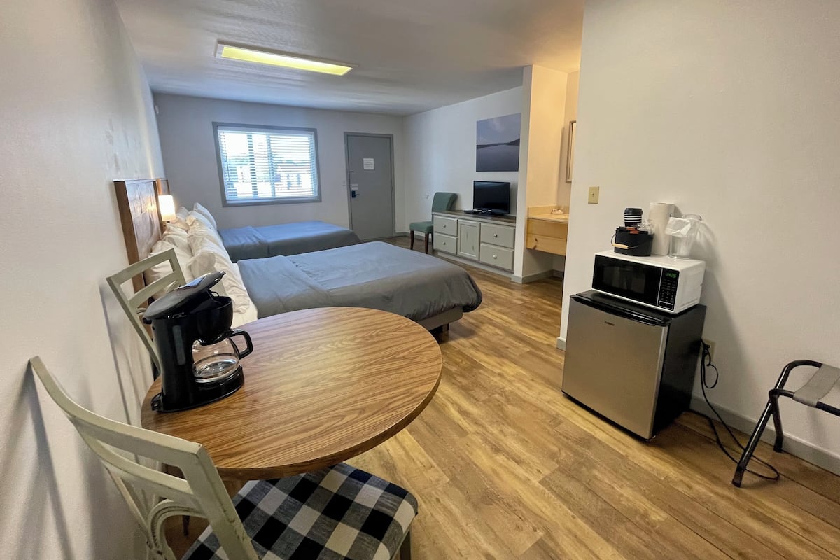 Quarto de hotel compacto com duas camas de solteiro, uma kitchenette pequena, um espaço de refeições com mesa e cadeiras, e uma televisão.