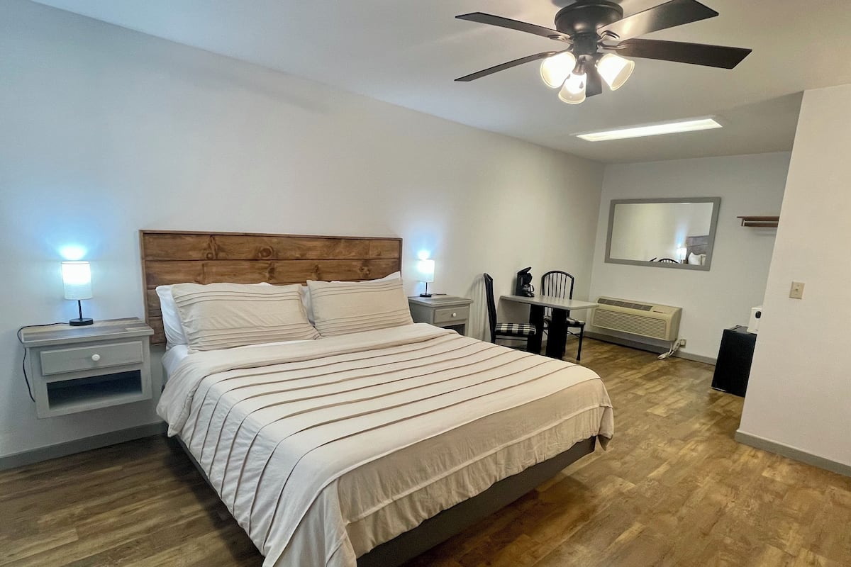 Quarto bem arrumado com cama, mesas de cabeceira, poltrona, escrivaninha, ventilador de teto e radiador.
