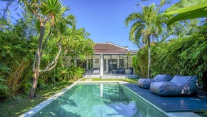 Piscina com cobertura de vidro, cercada por vegetação exuberante e uma casa moderna com telhado telhado.