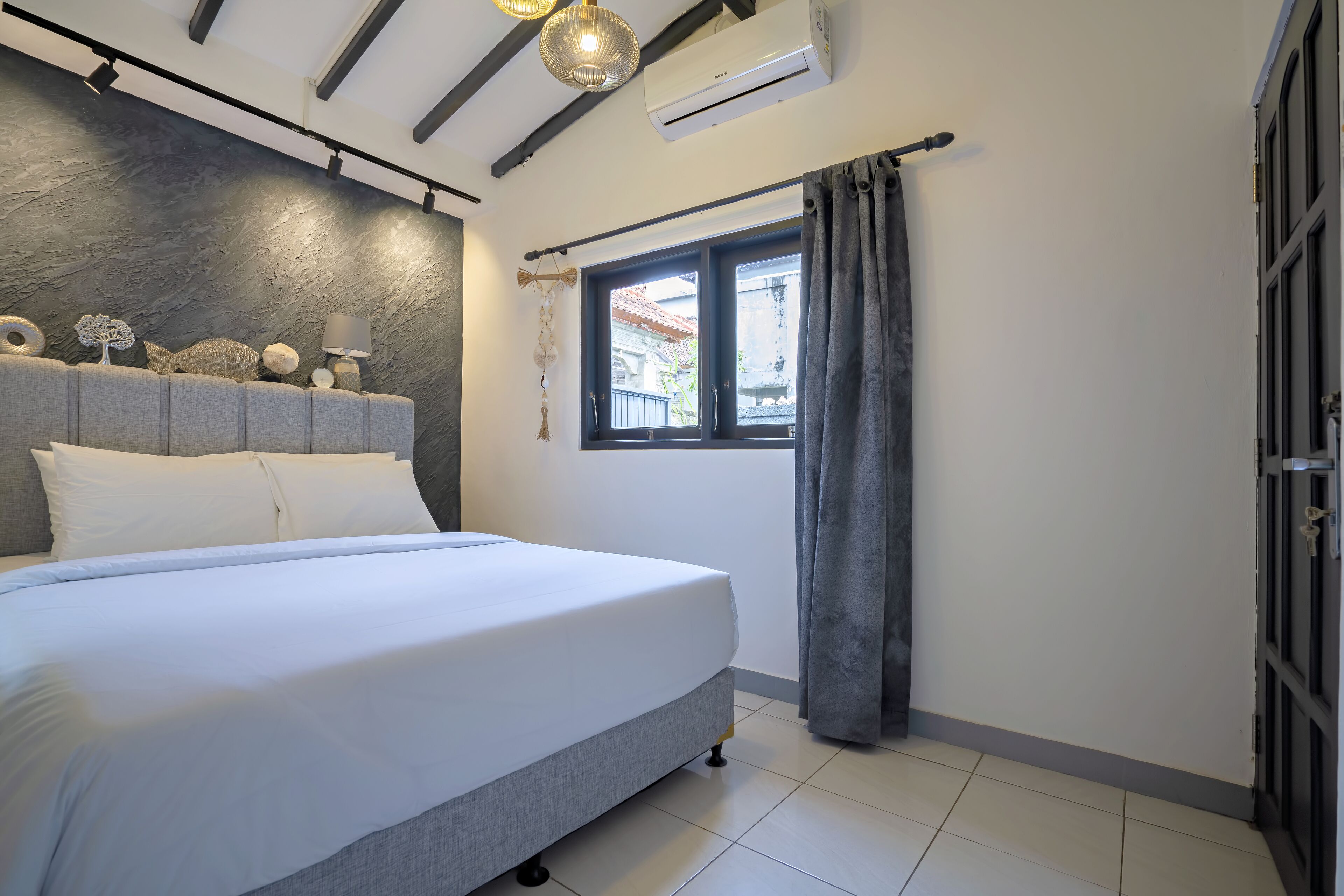 Quarto com cama, cabeceira, janela com cortinas, ar condicionado e luminária suspensa.