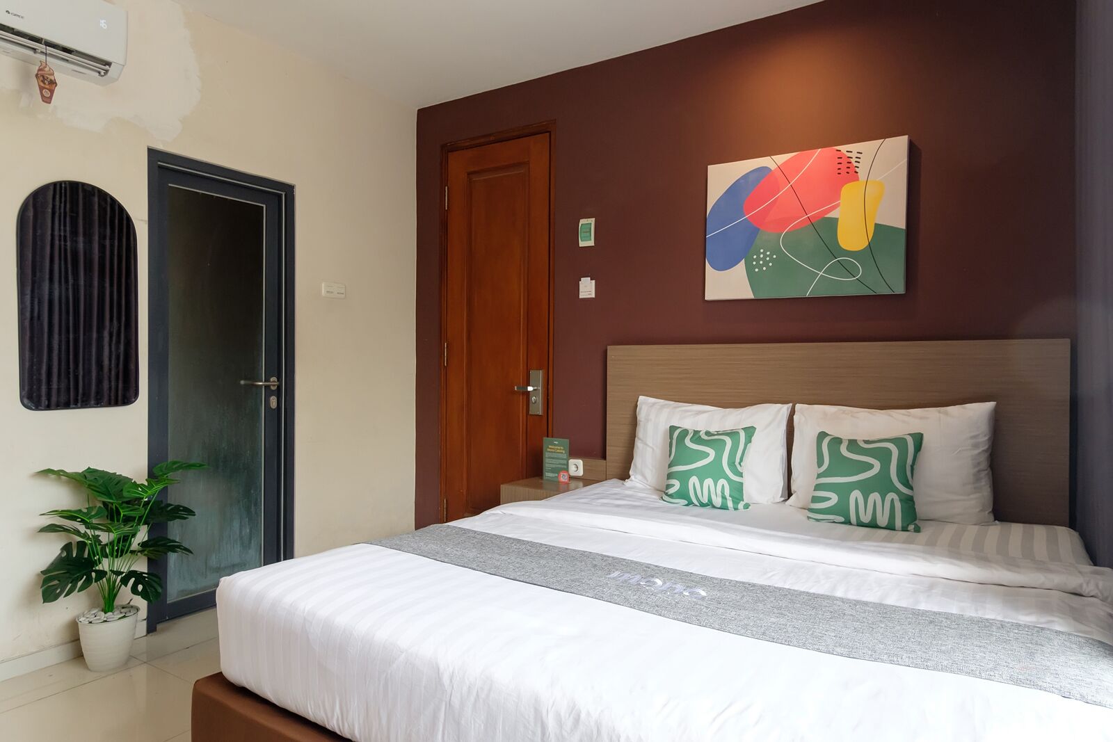 Quarto de hotel com cama, planta em vaso, porta e uma obra de arte na parede.