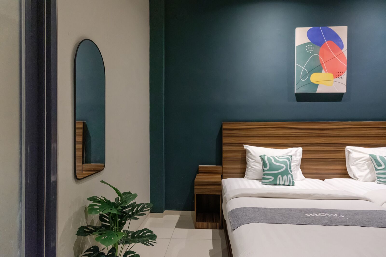 Quarto moderno com cabeceira de madeira, um espelho grande, mesa de cabeceira, planta em vaso e uma pintura abstrata colorida na parede.