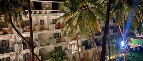 Um hotel com vários andares, varandas, palmeiras e uma área externa bem iluminada.