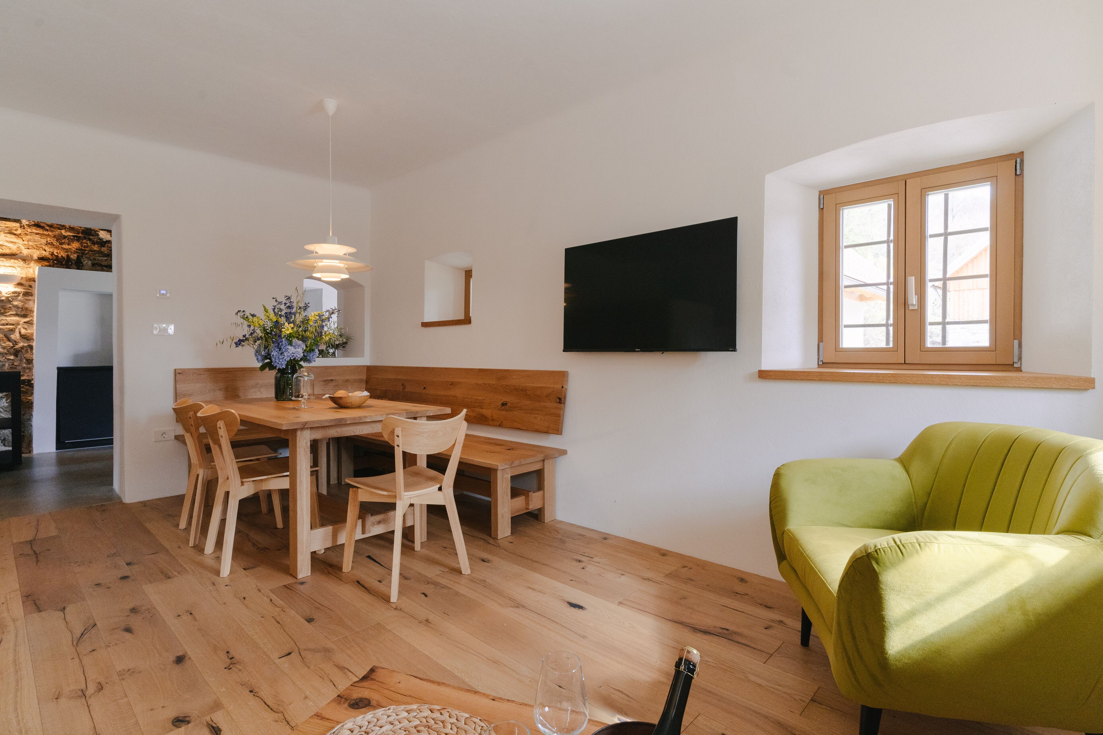 Uma sala de jantar com uma mesa e cadeiras de madeira, um sofá verde e uma TV fixada na parede.