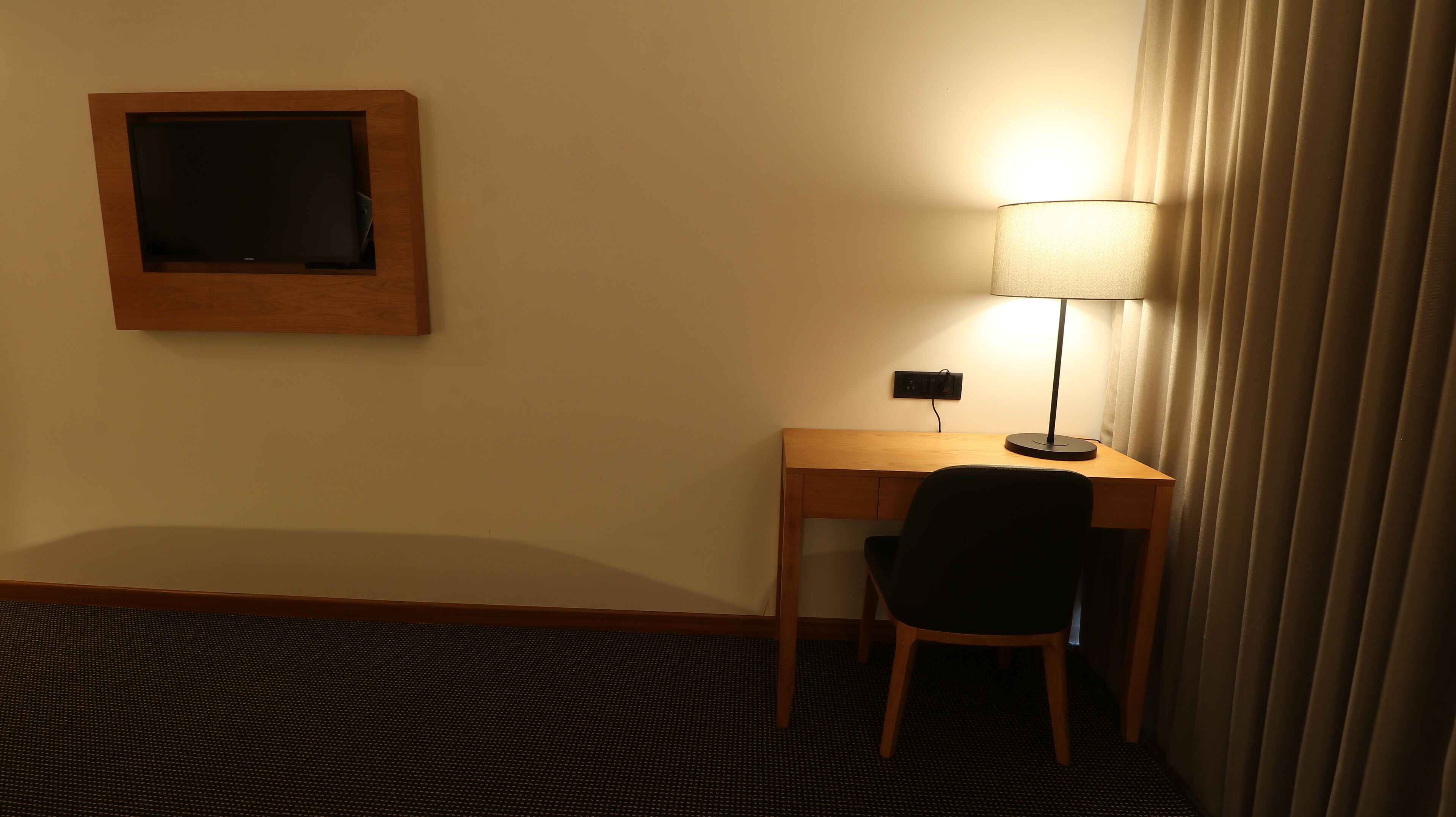 Quarto de hotel com escrivaninha de madeira, cadeira preta, luminária e quadro na parede.