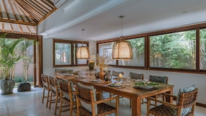 Uma sala de jantar espaçosa com uma mesa de madeira comprida, cadeiras de vime e grandes janelas com vista para o verde.