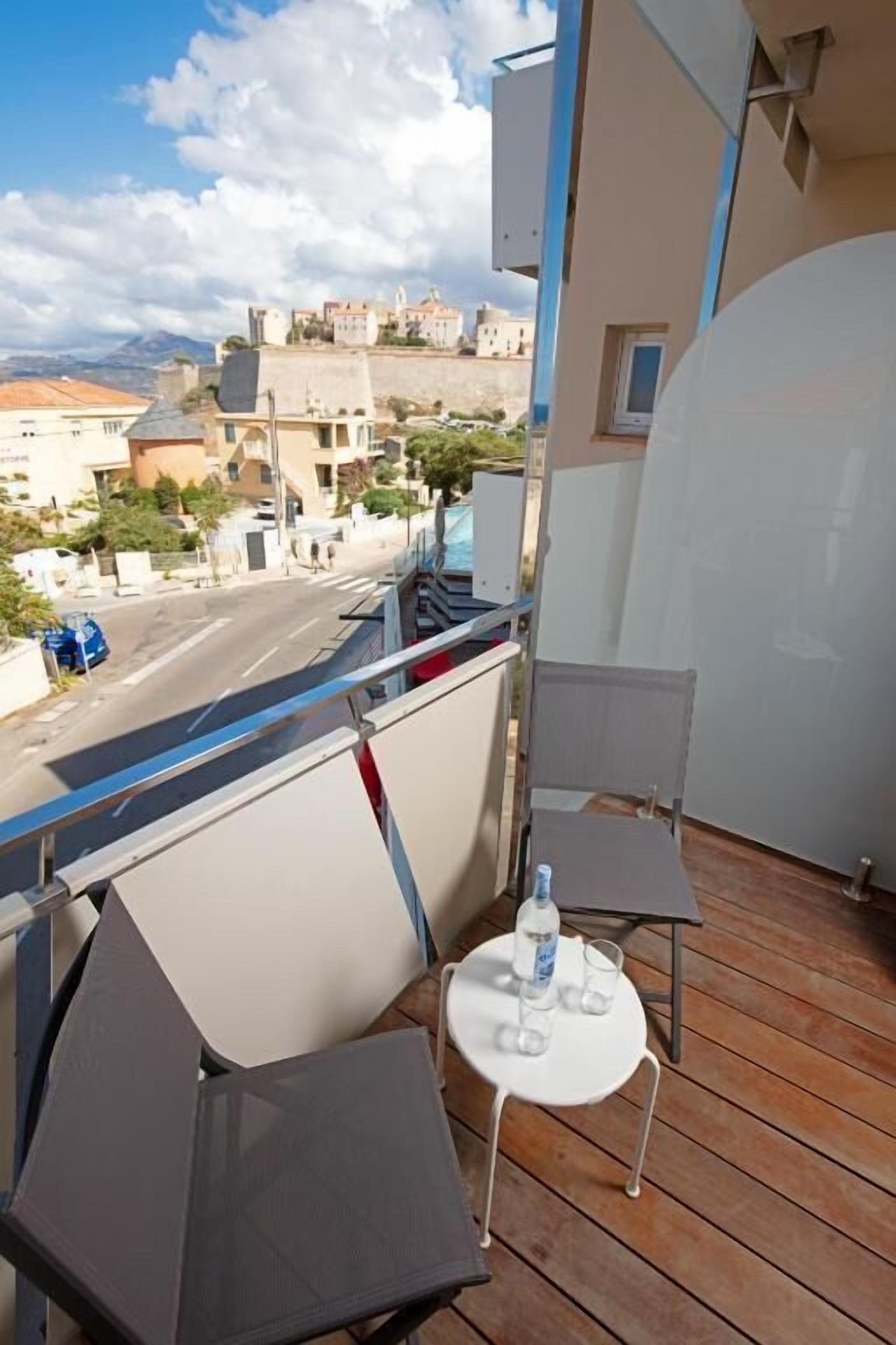 Uma varanda com vista para a cidade, uma mesa pequena com uma garrafa e copos, e duas cadeiras.