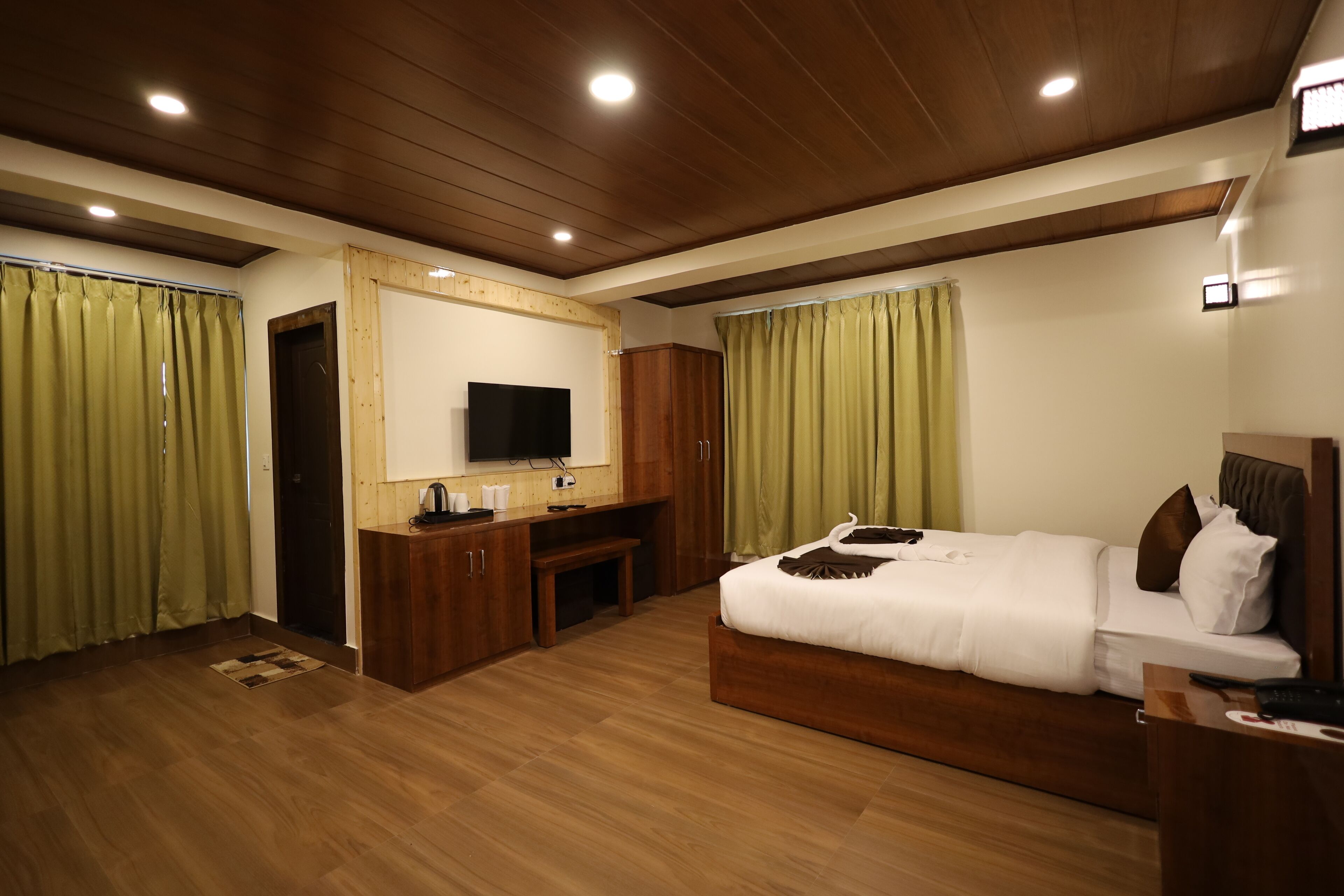 Quarto de hotel com teto de madeira, cama com roupa de cama branca, escrivaninha com televisão e cortinas verdes.