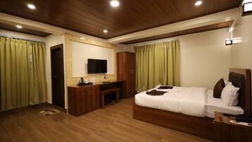 Quarto de hotel com teto de madeira, cama com roupa de cama branca, escrivaninha com televisão e cortinas verdes.