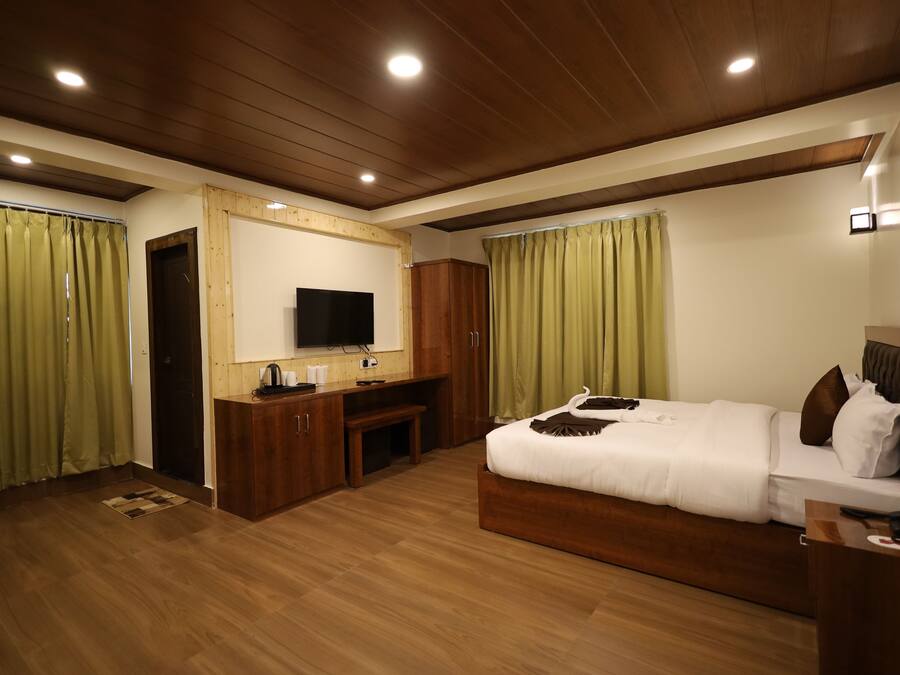 Quarto de hotel com teto de madeira, cama com roupa de cama branca, escrivaninha com televisão e cortinas verdes.