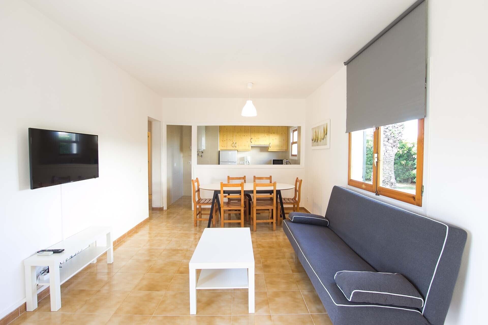 Uma sala de estar com televisão, sofá, mesa de jantar com cadeiras e cozinha ao fundo.