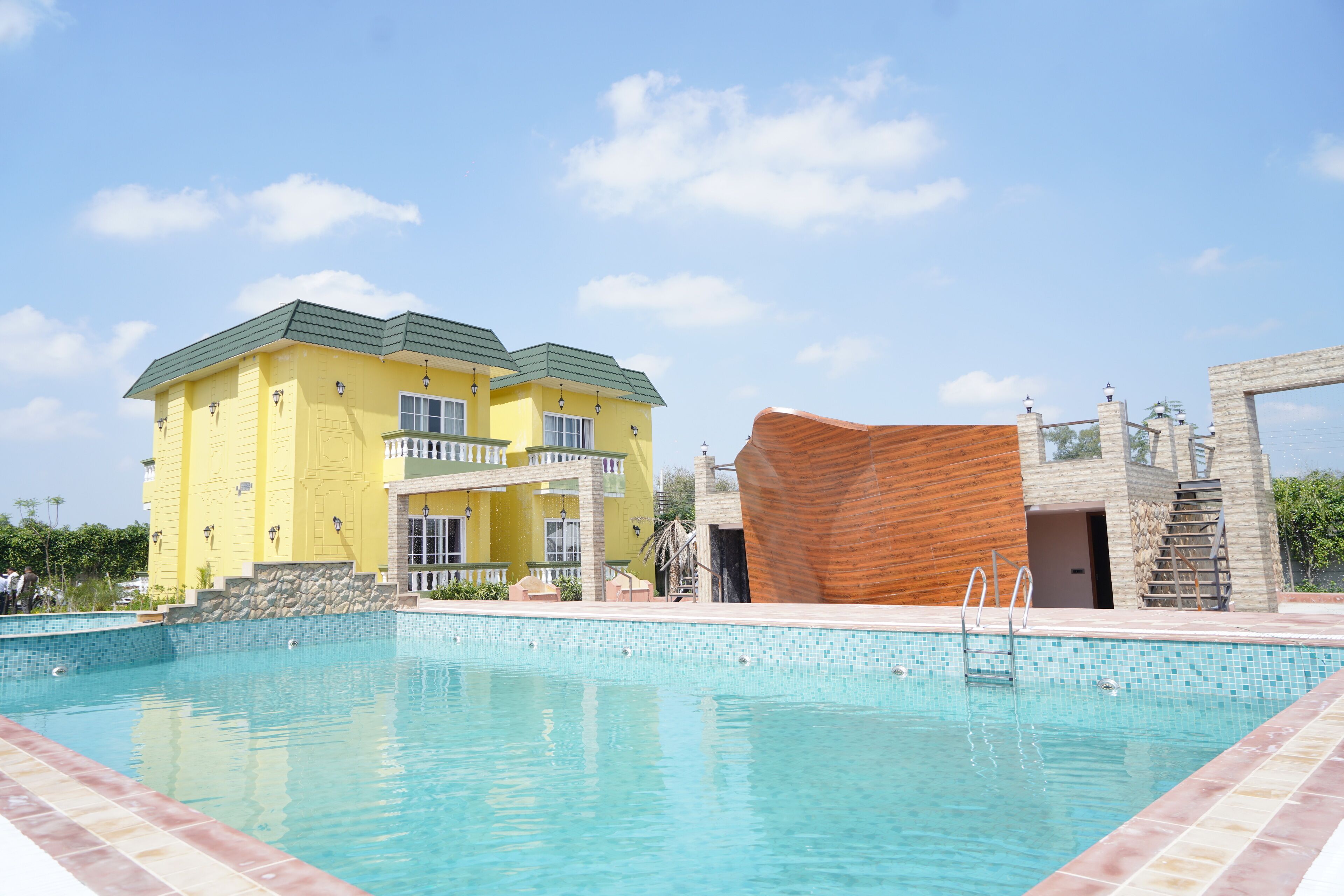 Uma piscina com borda azulejada, um edifício amarelo com telhado verde e uma estrutura de madeira com escadas.