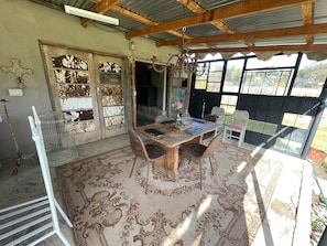 Uma área de refeições rústica, com uma mesa e cadeiras de madeira, um lustre de metal decorativo e vista para o exterior através de janelas amplas.