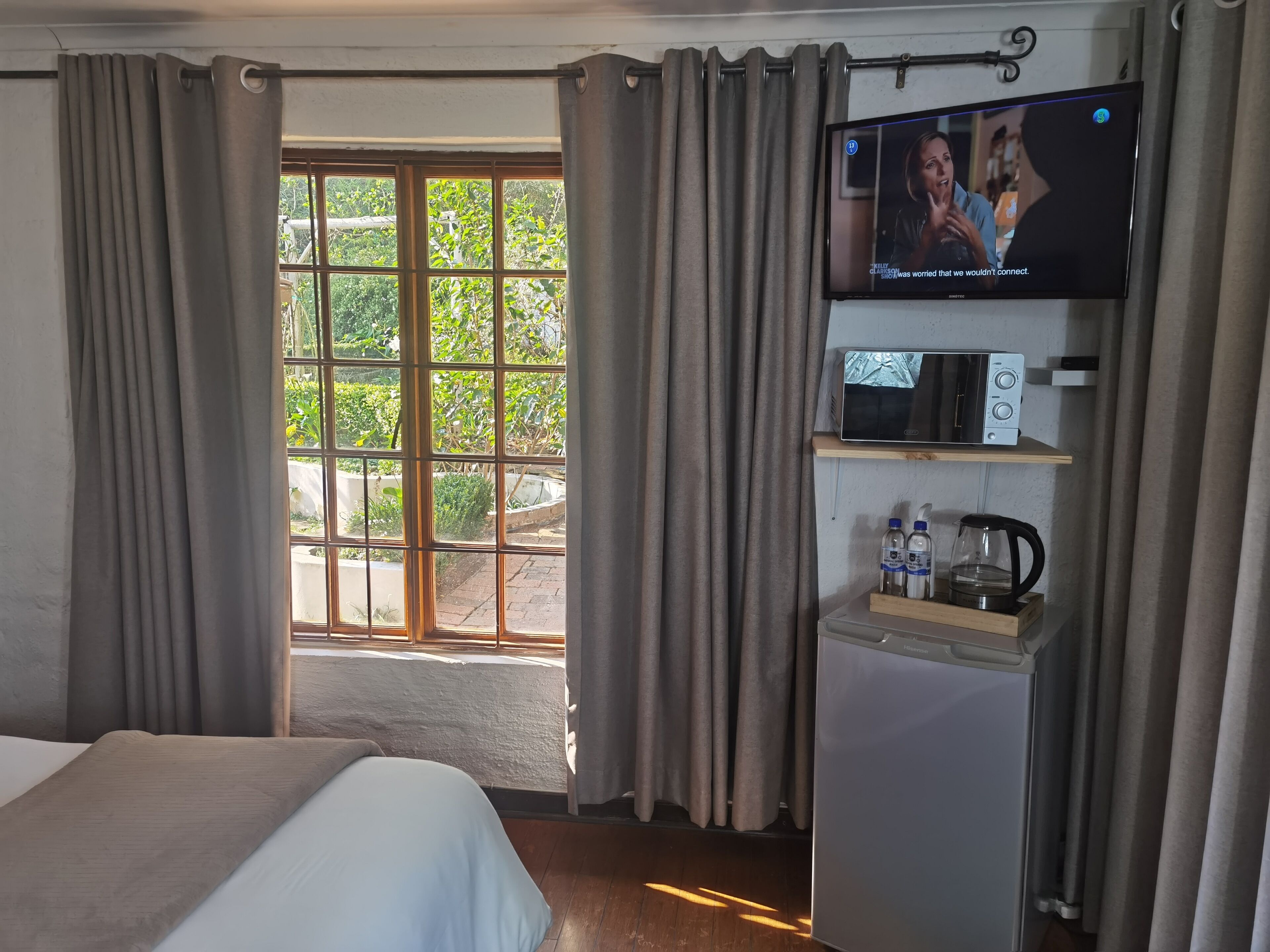 Quarto de hotel com cama, televisão em suporte, micro-ondas e uma pequena prateleira com garrafas e chaleira.