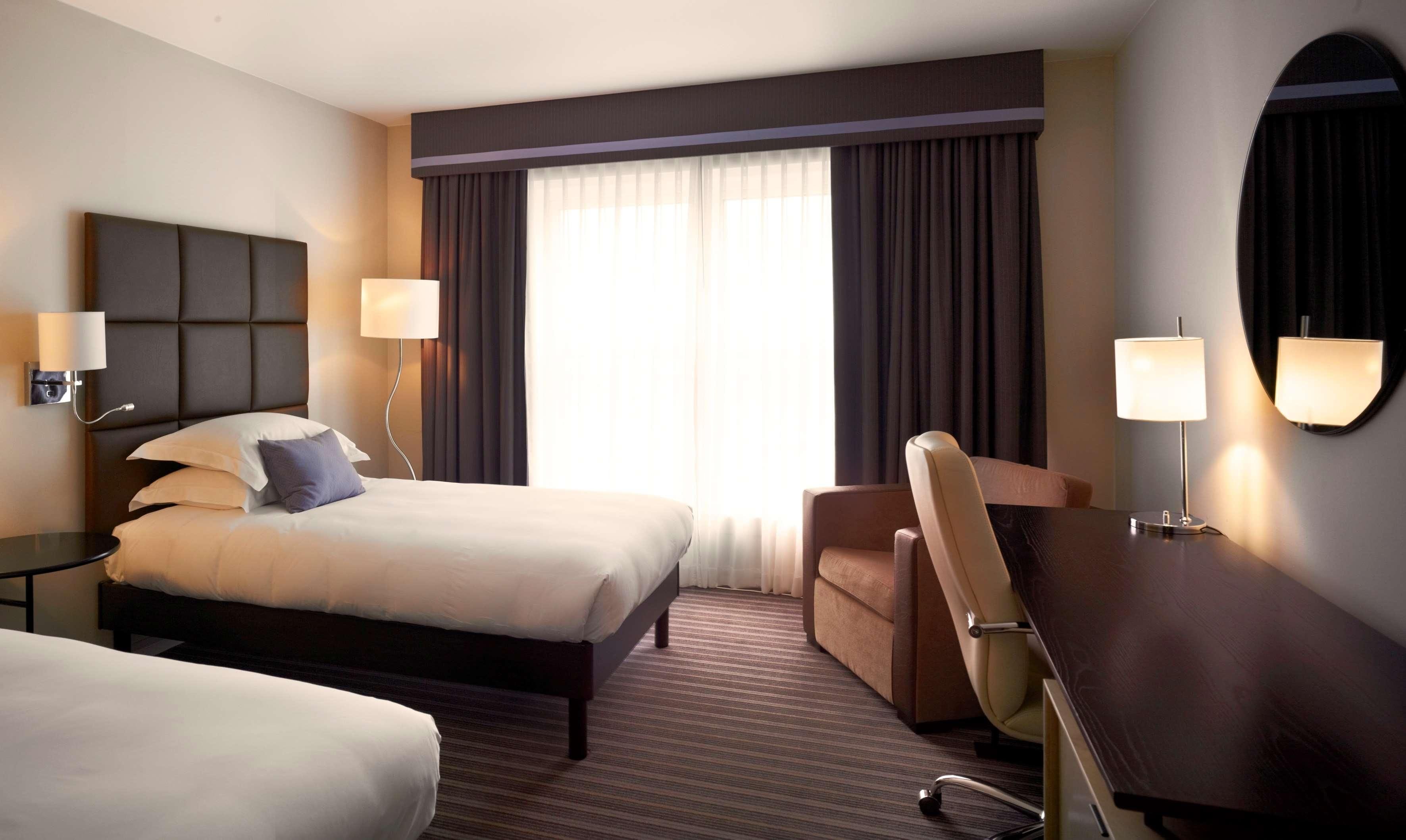 Quarto de hotel com uma cama grande, uma escrivaninha, duas luminárias, uma cadeira e uma janela ampla com cortinas.