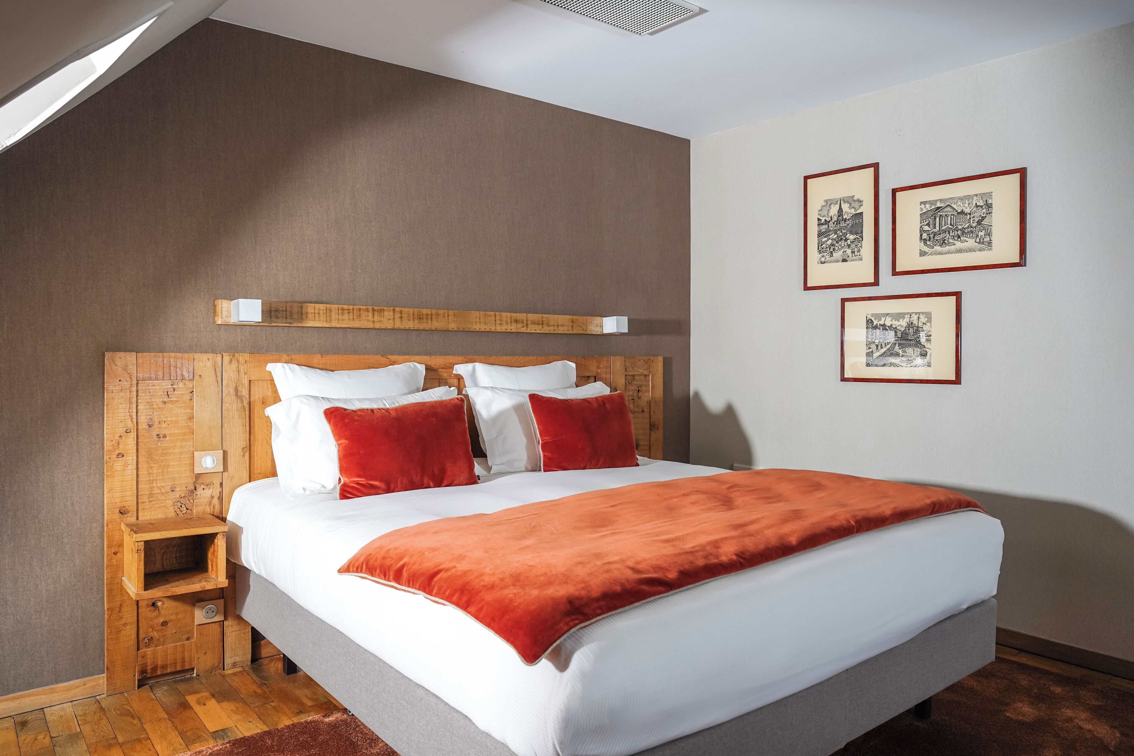 Quarto com cabeceira de madeira, roupa de cama branca e manta vermelha. Há três quadros na parede.
