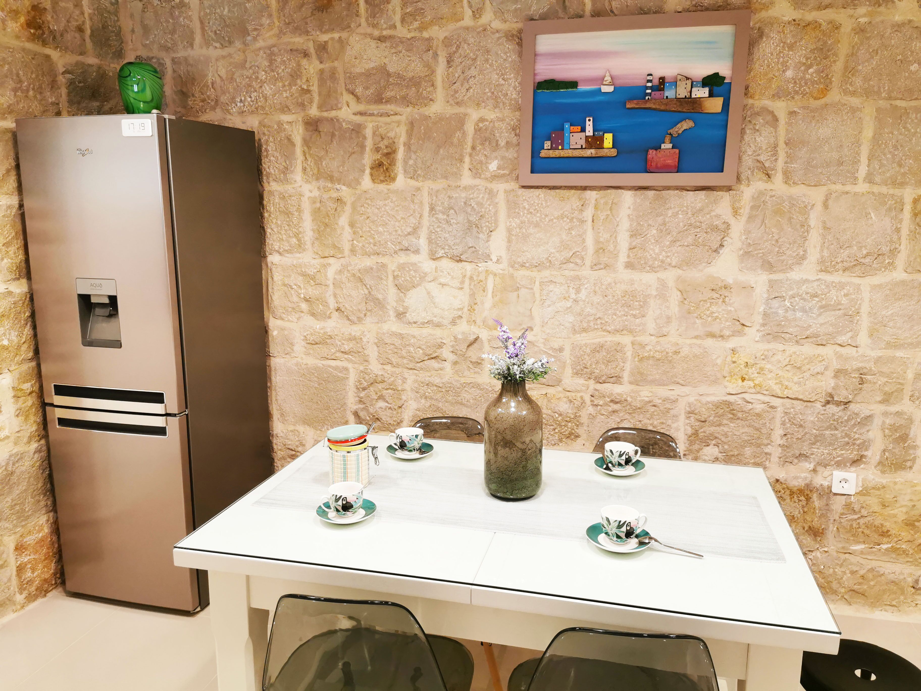 Uma cozinha com parede de pedra, geladeira, mesa com um vaso de flores e um quadro na parede.