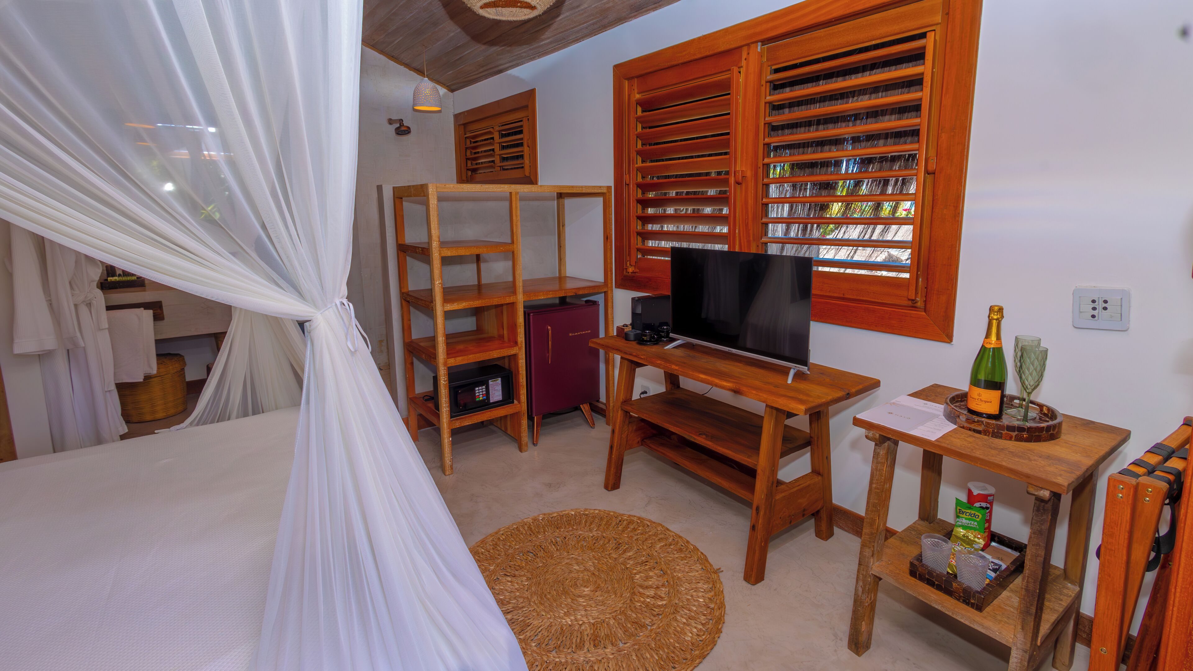 Quarto com cama, televisão, uma mesinha com garrafa e copos, e prateleiras de madeira.