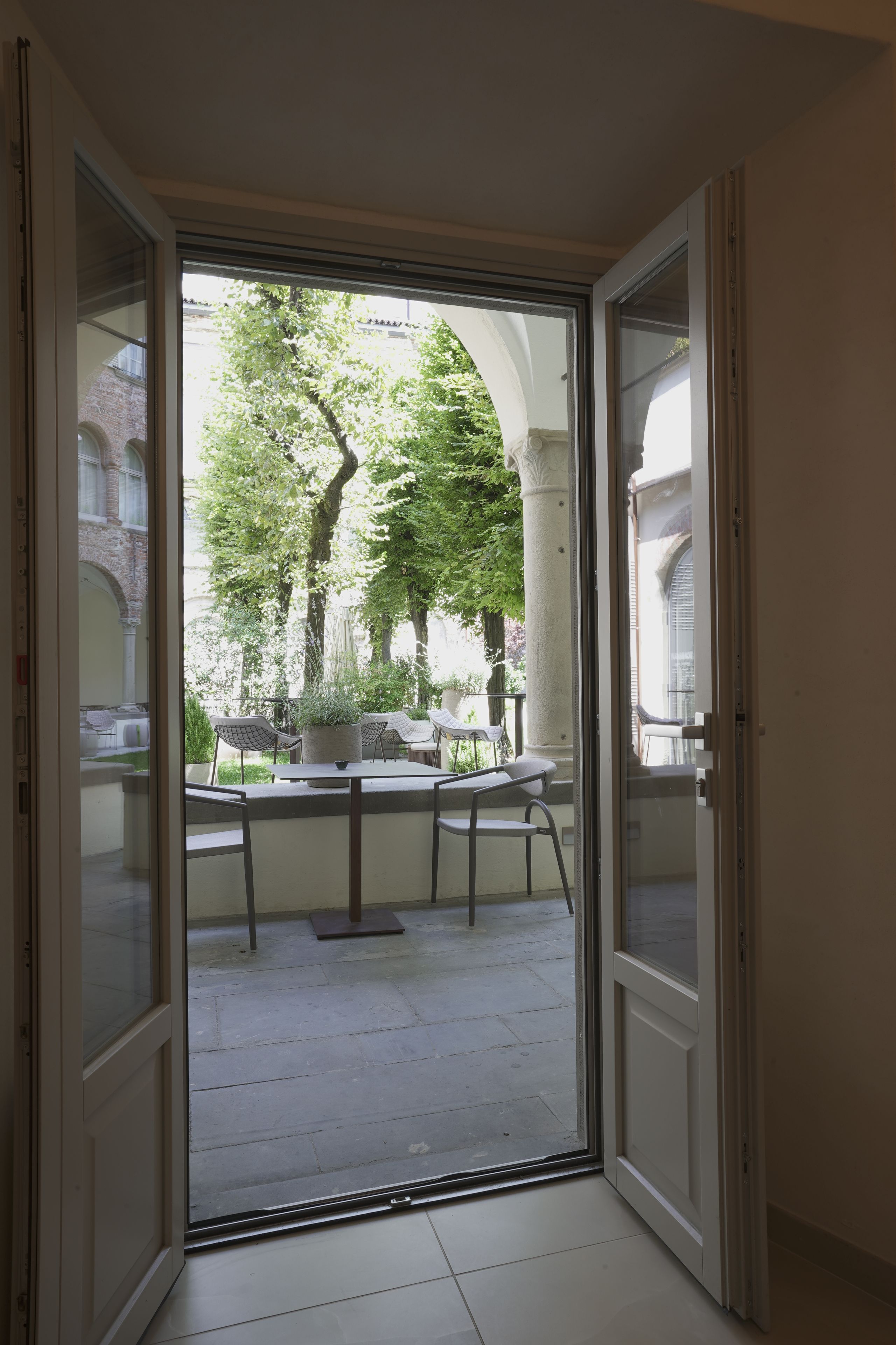 Visão através de uma porta de vidro para um pátio com mesa e cadeiras, rodeado por árvores e um edifício de tijolos.