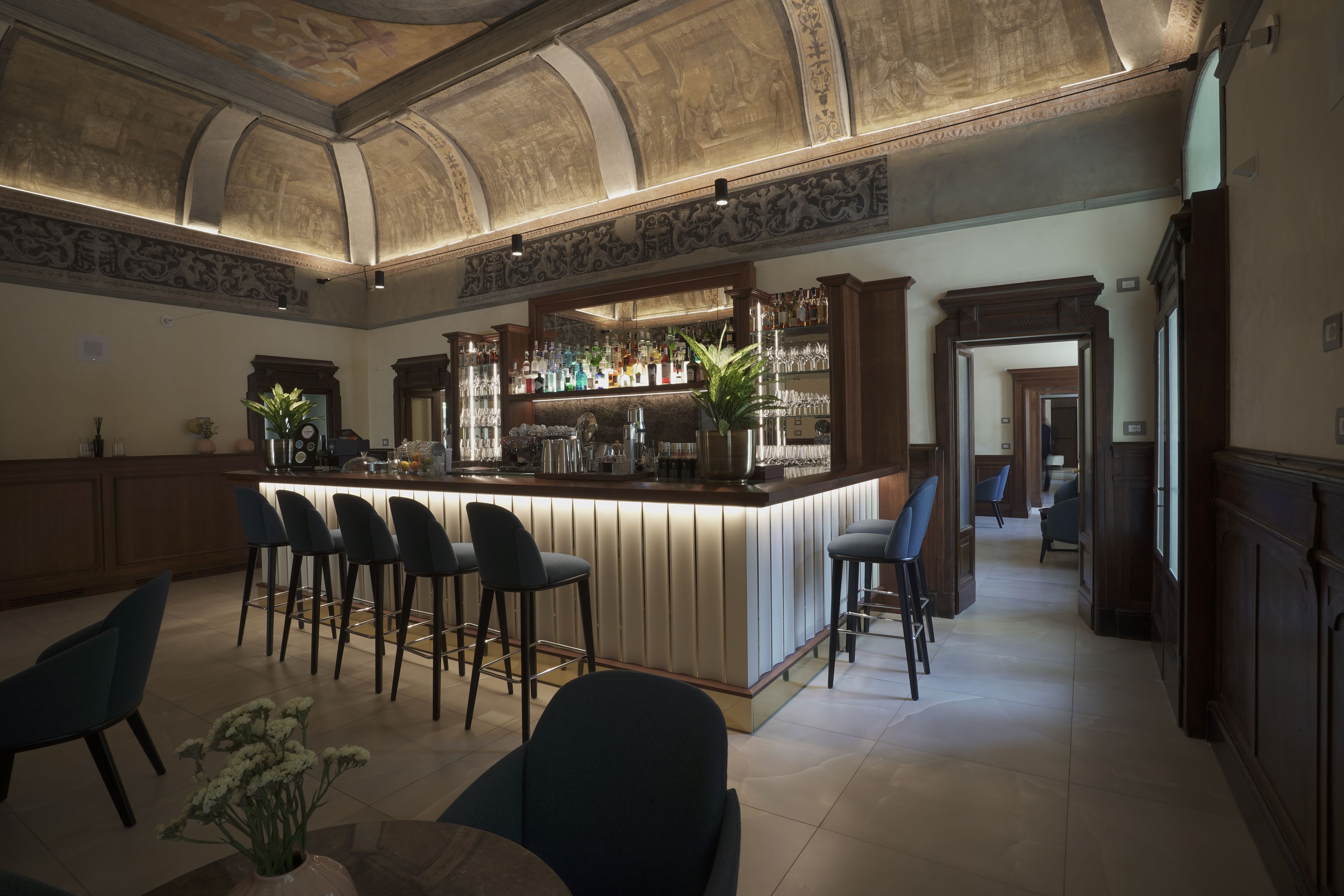 Uma área de bar com teto alto e design ornamentado, banquetas e um balcão com diversos copos e garrafas.
