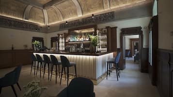 Uma área de bar com teto alto e design ornamentado, banquetas e um balcão com diversos copos e garrafas.