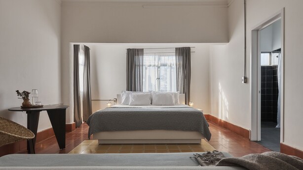 Quarto de hotel moderno com uma cama grande, uma mesa pequena com um vaso e vista para a cidade pela janela.