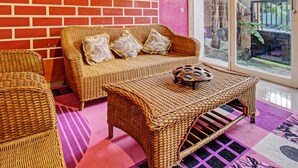 Sofá de vime com almofadas, uma poltrona combinando e uma mesa de centro de fibra natural num espaço com parede de tijolos e porta de vidro que dá para o exterior.