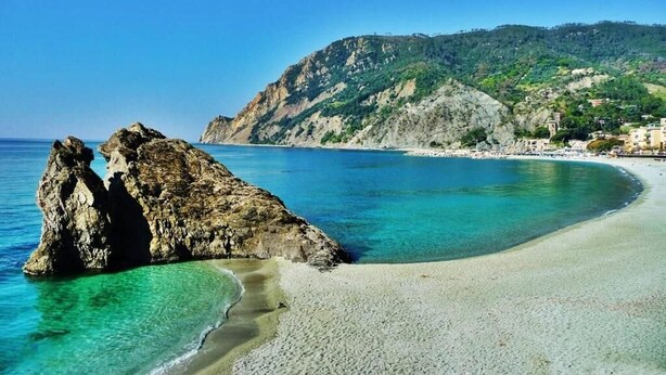 Uma praia com águas turquesa cristalinas, um afloramento rochoso e um morro com edifícios ao fundo.