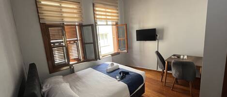 Quarto com cama, escrivaninha, cadeira, televisão e janela com persianas.
