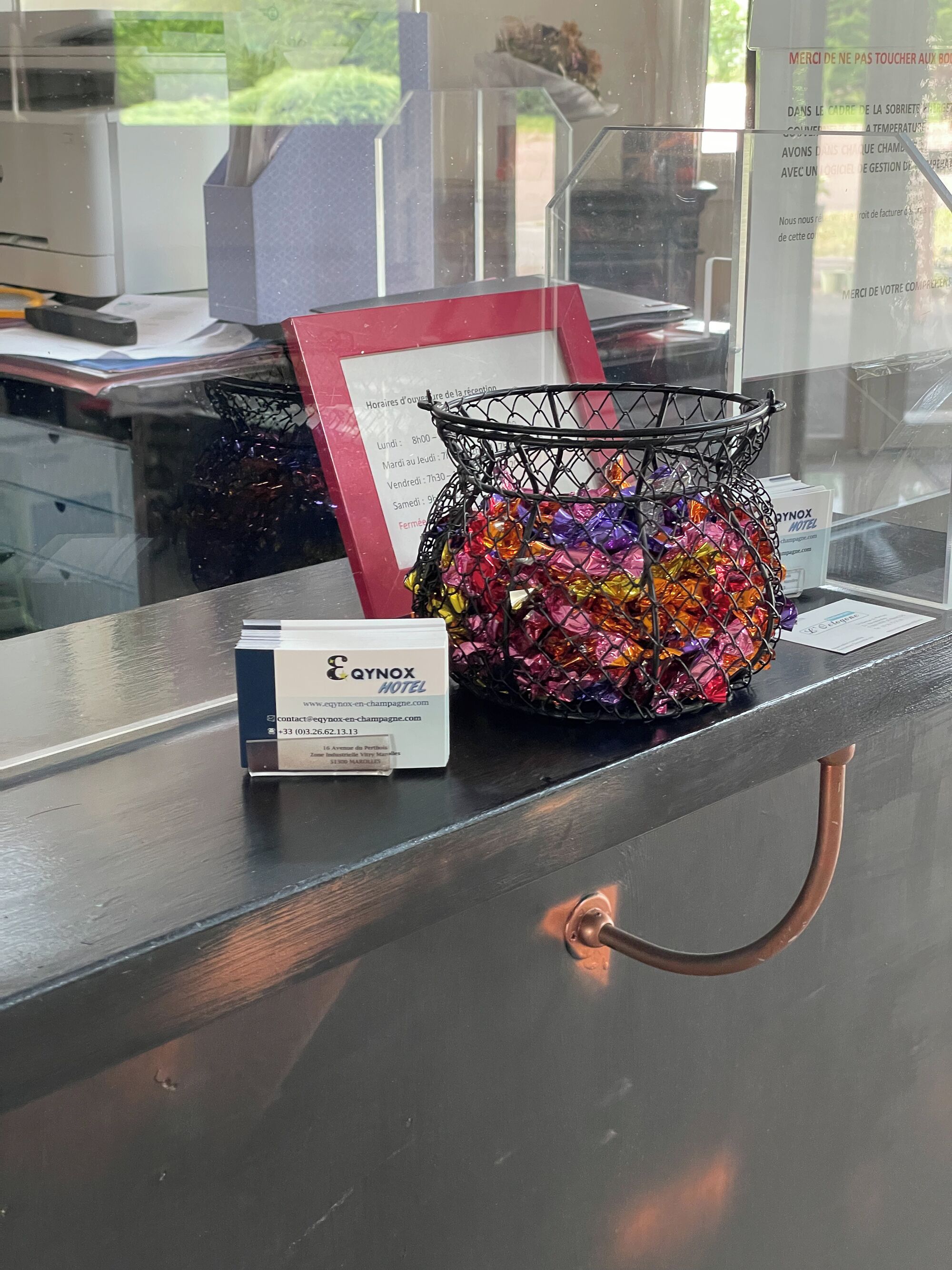 Uma área de recepção de hotel com uma vitrine de vidro contendo itens coloridos embalados e um aviso com informações de contato do hotel.