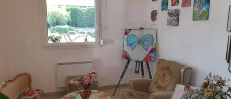 Um quarto acolhedor com uma mesa redonda, sofá floral e uma cadeira. Uma janela grande oferece vista para o verde lá fora.