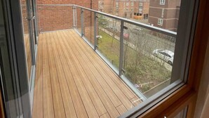 Varanda com piso de madeira e guarda-corpo de metal, com vista para um edifício de tijolos e uma área gramada.