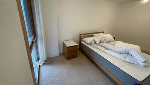 Quarto pequeno com uma cama de solteiro, cabeceira de madeira, mesa de cabeceira e janela com cortinas.